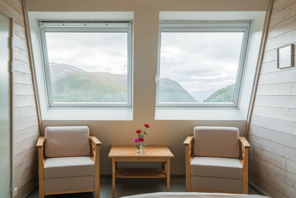 Quarto com duas janelas com vista para as montanhas, uma pequena mesa com um vaso de flores e duas cadeiras.