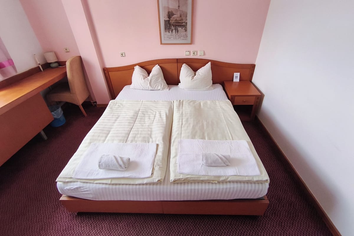 Quarto de hotel com uma cama de casal, duas almofadas, cabeceira de madeira, escrivaninha, cadeira, mesa de cabeceira e um quadro pendurado na parede.