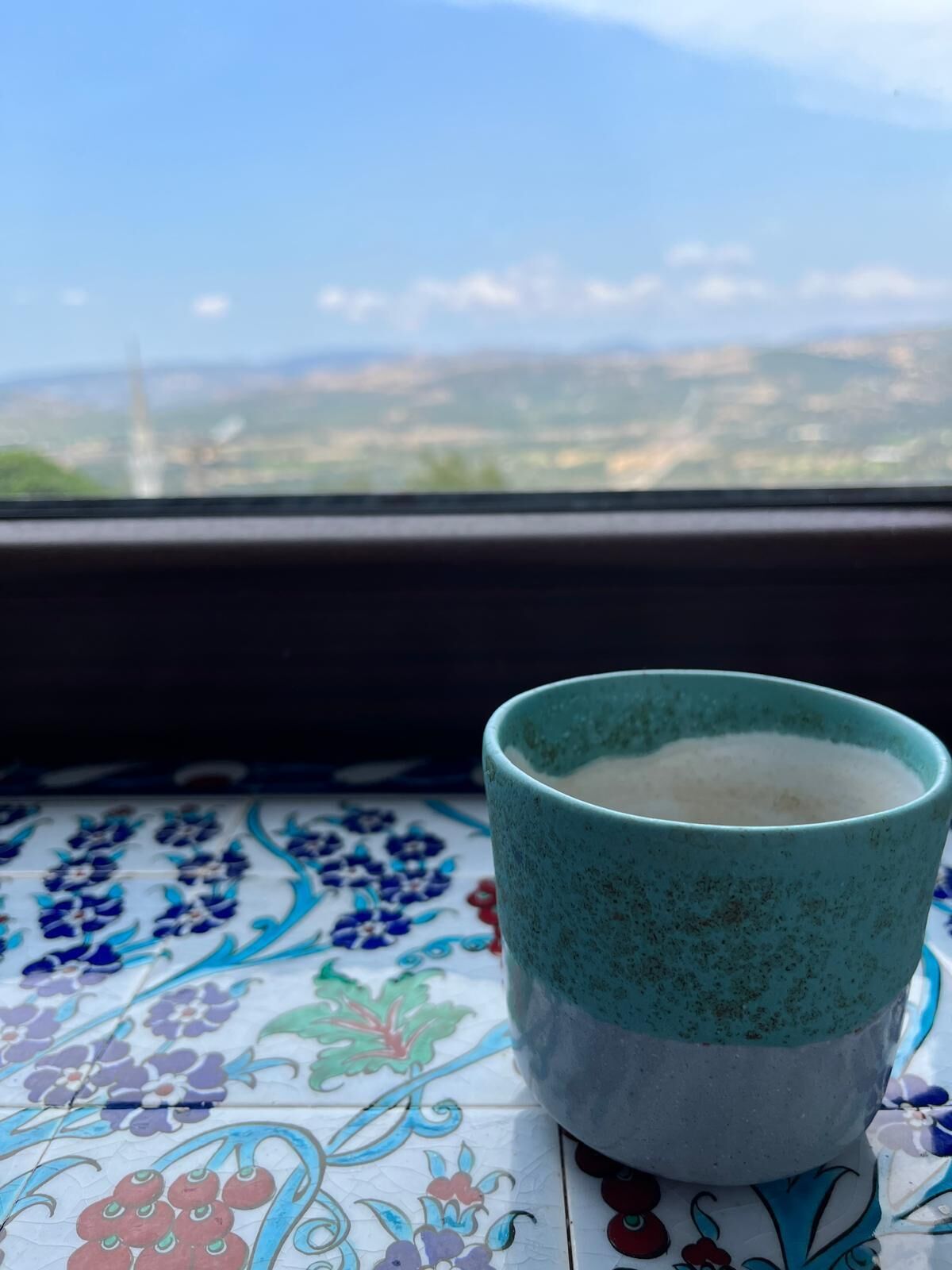 Uma caneca azul-esverdeada sobre uma toalha de mesa estampada, perto de uma janela com vista para uma paisagem distante.