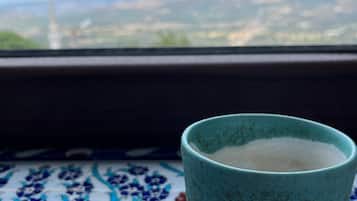 Uma caneca azul-esverdeada sobre uma toalha de mesa estampada, perto de uma janela com vista para uma paisagem distante.
