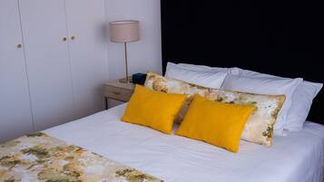 Cama com roupa de cama branca e duas almofadas amarelas, mesa de cabeceira com abajur e criado-mudo com um objeto azul.