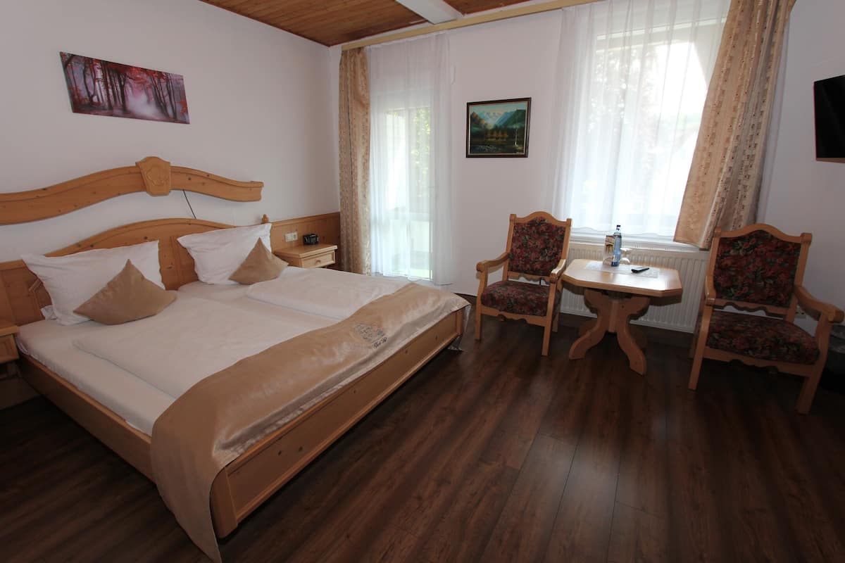 Quarto com cama de madeira, duas cadeiras, uma mesinha e janela com cortinas.