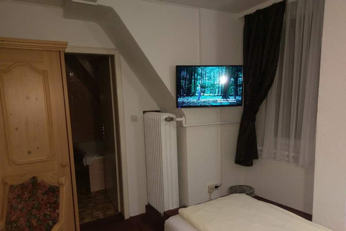 Quarto com uma cama, televisão fixada na parede, radiador e porta que dá para outro espaço.