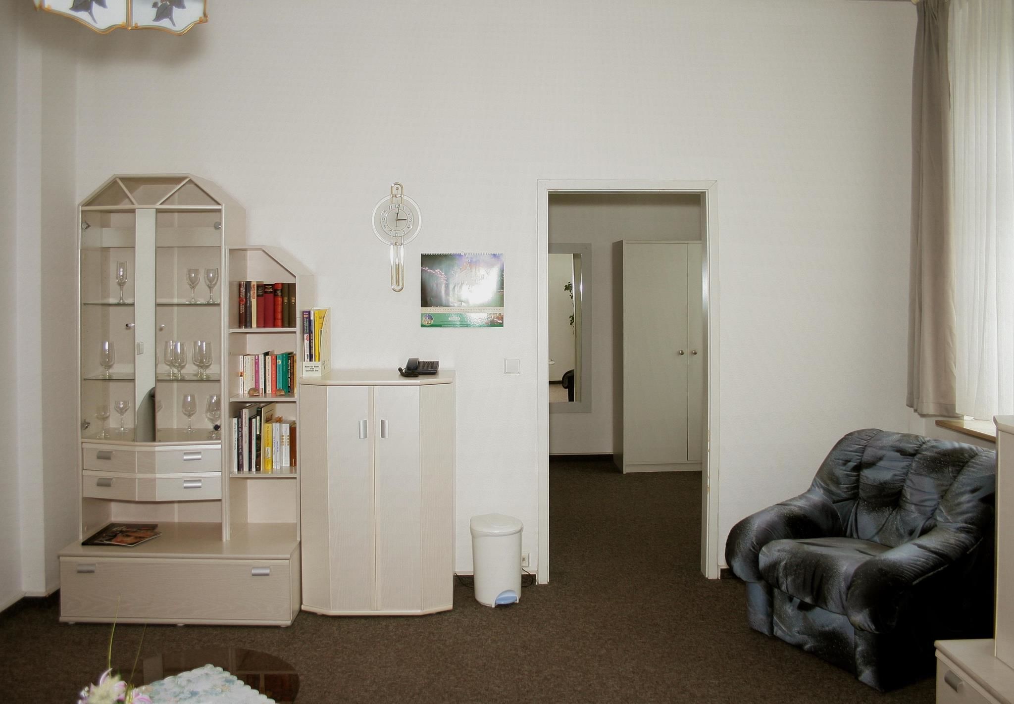 Uma sala de estar com uma estante de livros, uma poltrona de couro preto, uma mesa pequena com um vaso e uma porta que dá para outro cômodo.