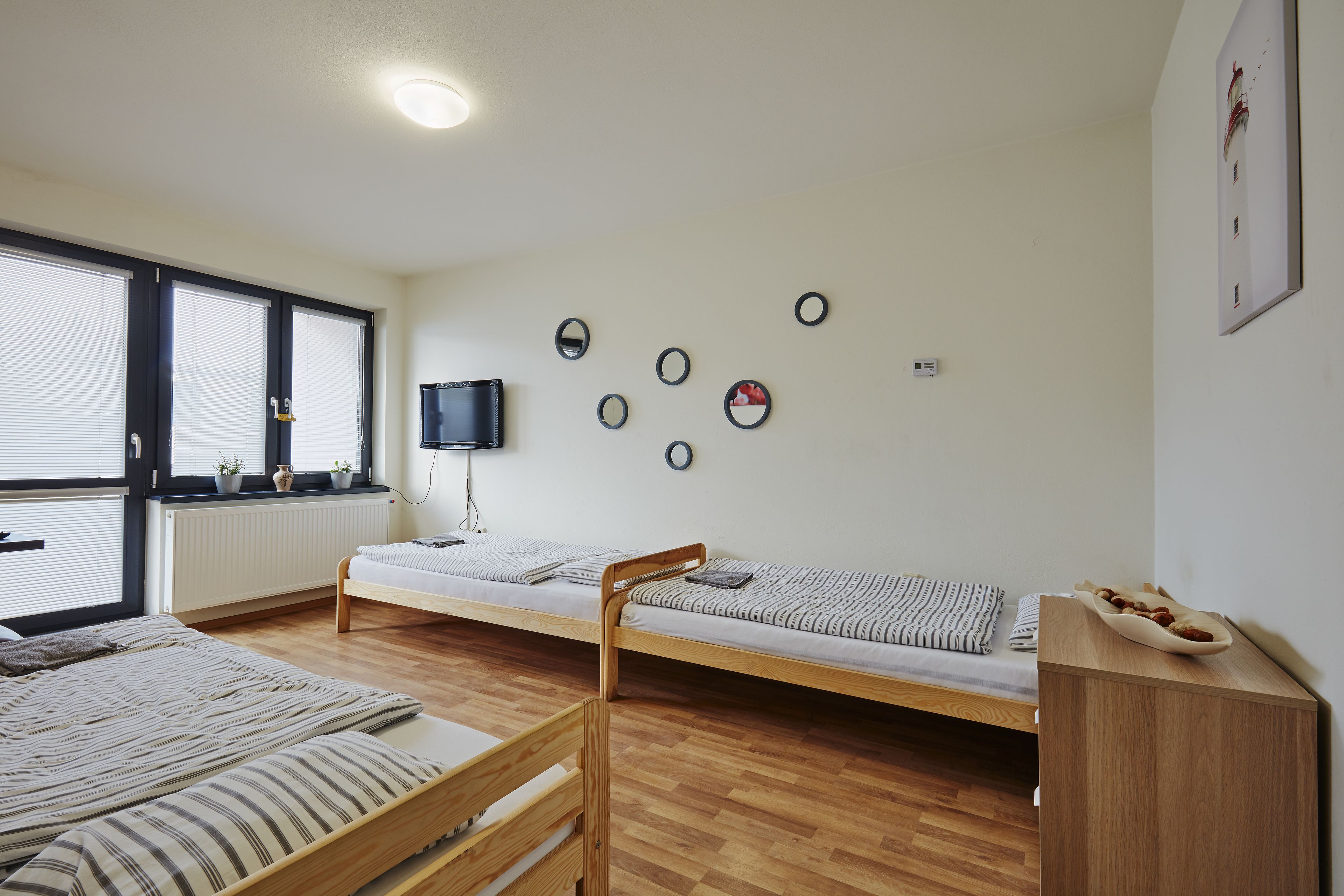 Quarto com duas camas de solteiro, piso de madeira, televisão na parede e janela com persianas.