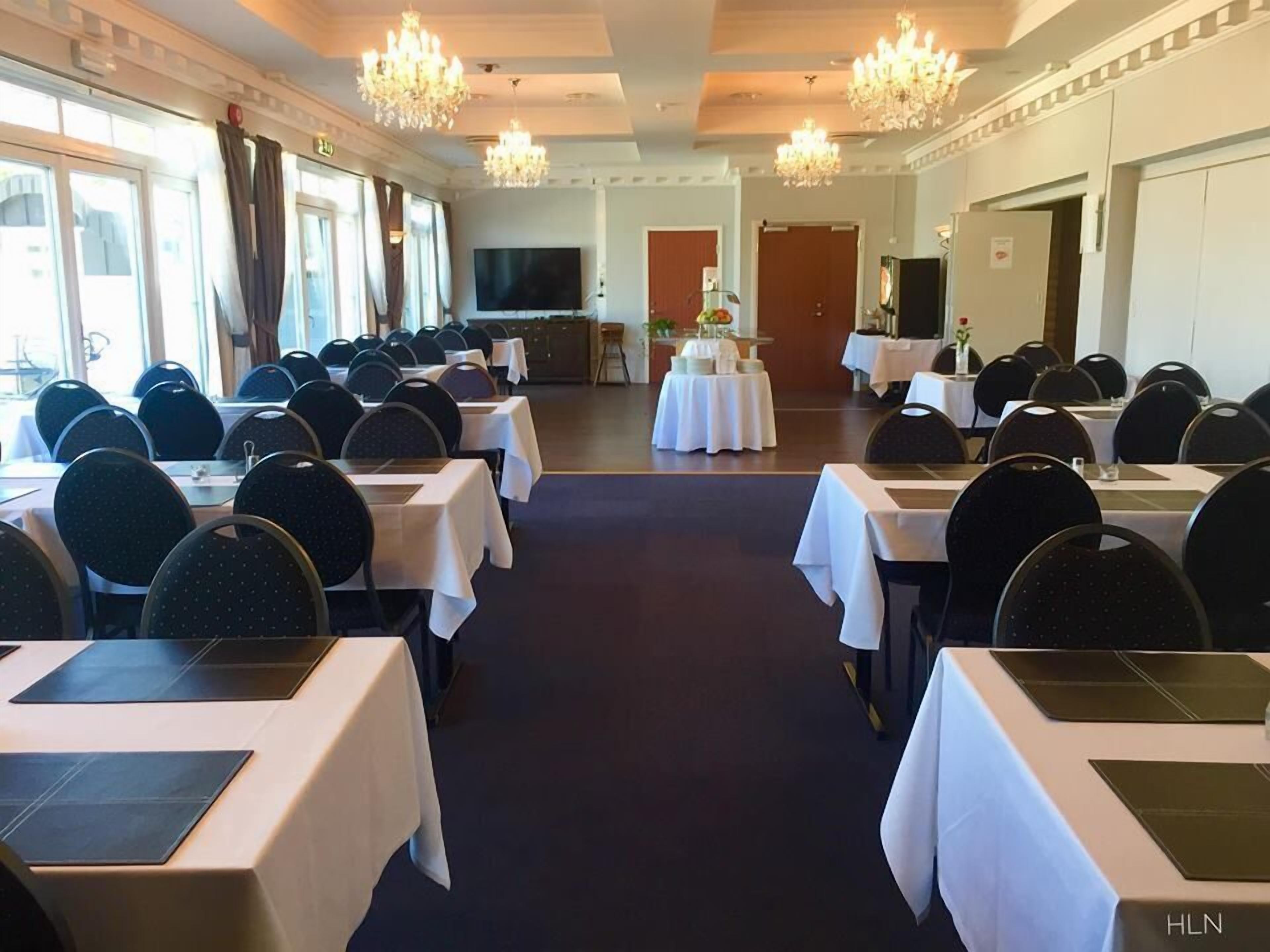 Uma sala de conferências com mesas, cadeiras, uma tela grande e lustres.