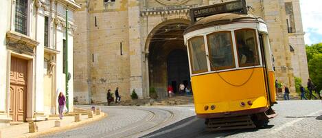 Um edifício histórico com uma grande entrada em arco e duas torres. Um elétrico amarelo passa na frente.
