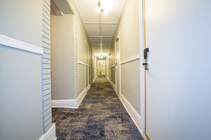 Corredor de hotel com piso carpetado, paredes brancas e uma porta à direita.