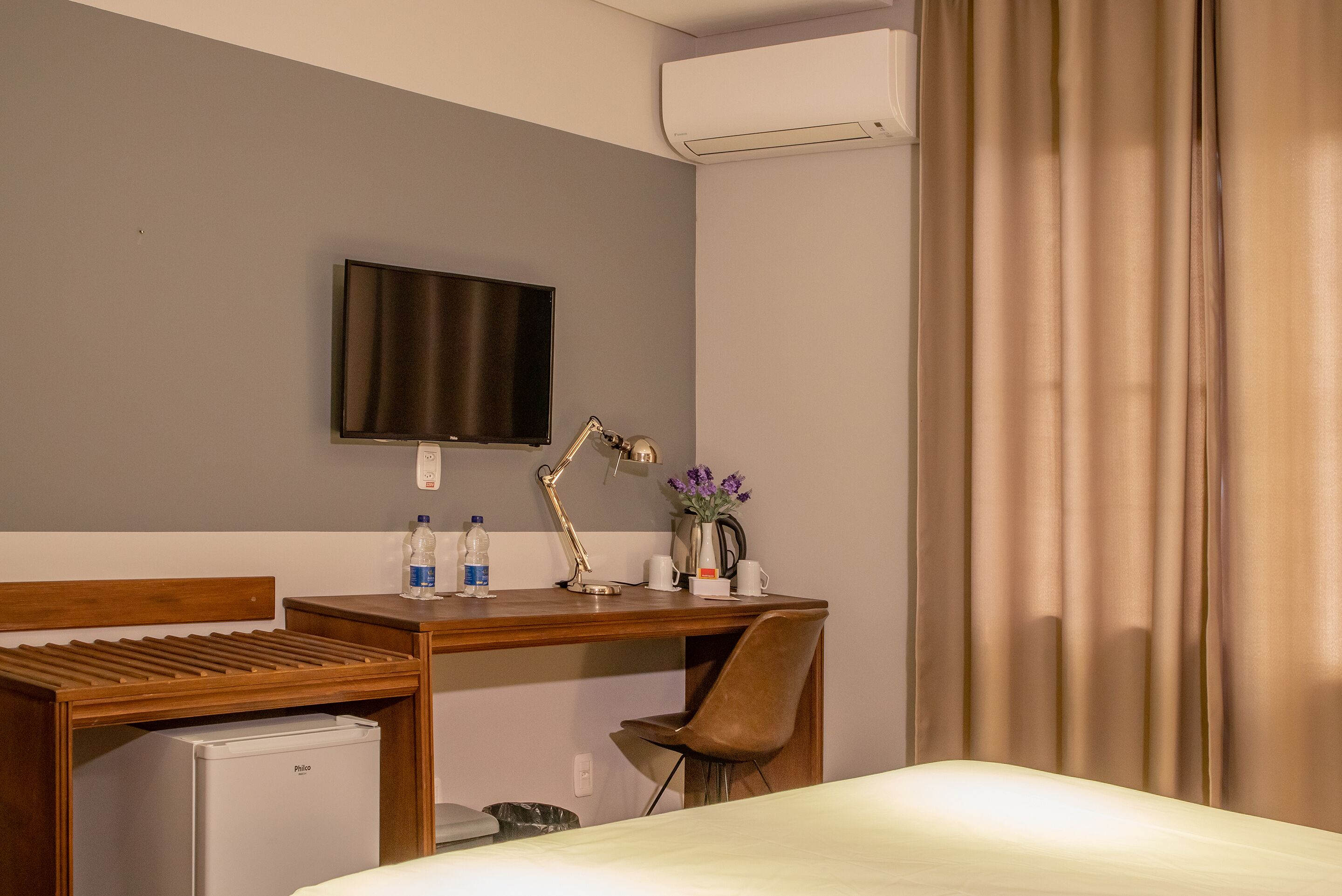 Quarto de hotel com uma escrivaninha de madeira, um pequeno frigorífico, uma TV na parede, uma cadeira e uma cama.
