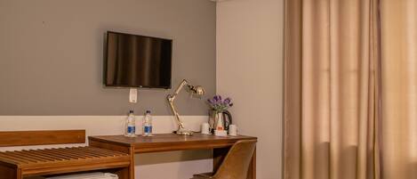 Quarto de hotel com uma escrivaninha de madeira, um pequeno frigorífico, uma TV na parede, uma cadeira e uma cama.