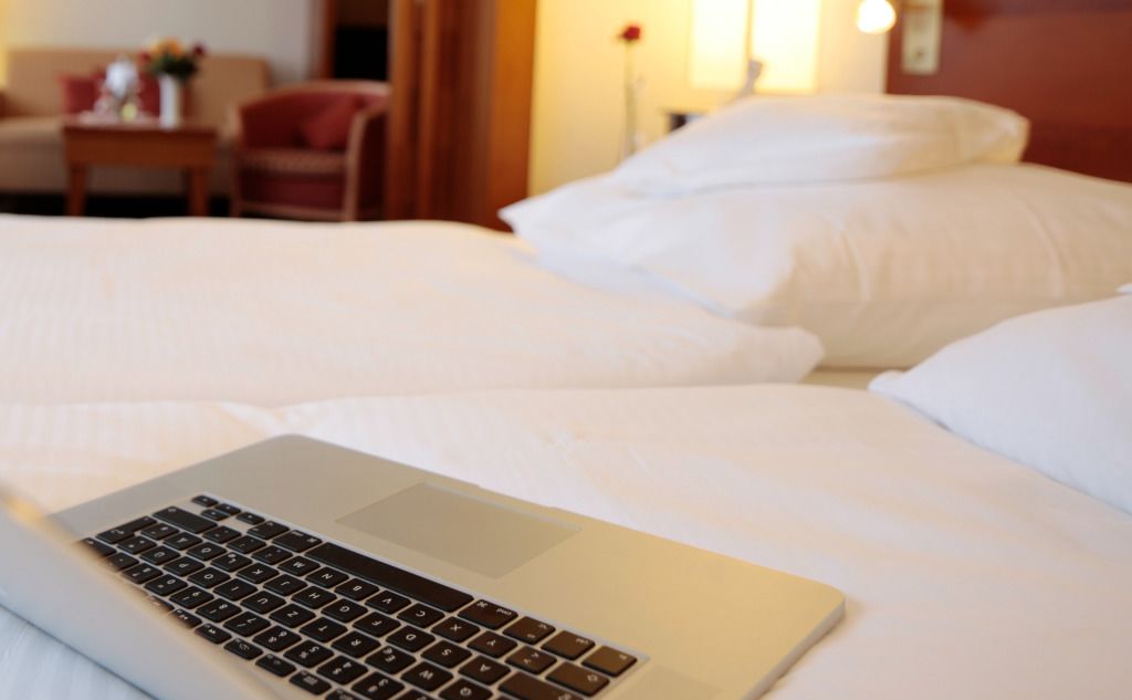 Quarto de hotel com uma cama, um laptop sobre a cama e uma mesa de cabeceira com uma luminária.