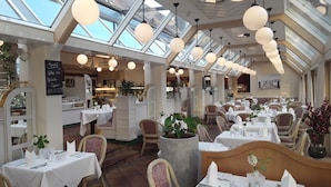 Restaurante com pé-direito alto, claraboias e luminárias pendentes redondas. Mesas dispostas com toalhas brancas e arranjos de mesa.
