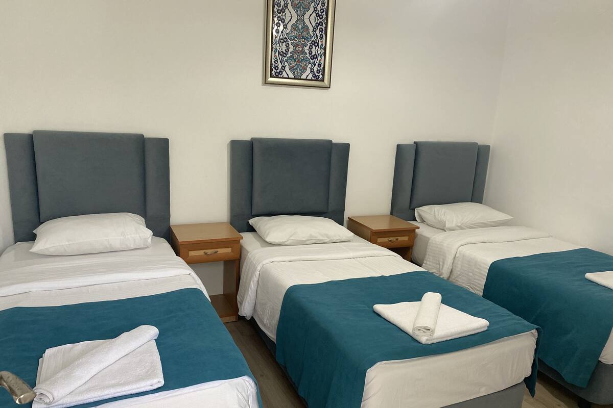 Quarto de hotel com quatro camas de solteiro, cada uma com cabeceira cinza, roupa de cama branca e manta azul-turquesa. Há uma mesa de cabeceira de madeira com gaveta entre cada cama, e uma obra de arte emoldurada acima das cabeceiras.