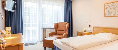 Quarto de hotel com cama, escrivaninha com telefone, cadeira, televisão presa na parede e janela com cortinas.