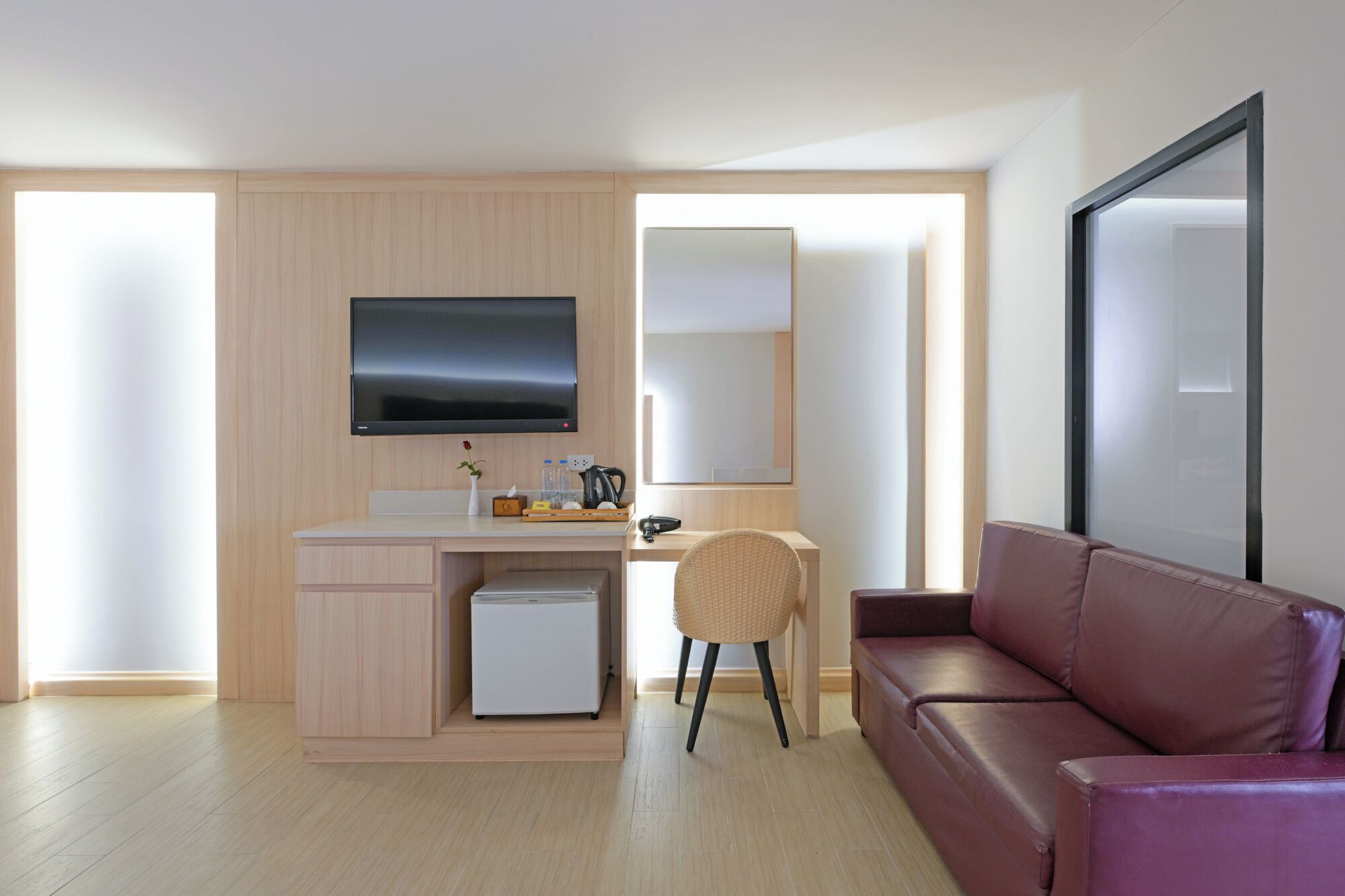 Quarto de hotel moderno com escrivaninha, televisão, espelho, pequeno frigorífico, cadeira de vime e sofá de couro.