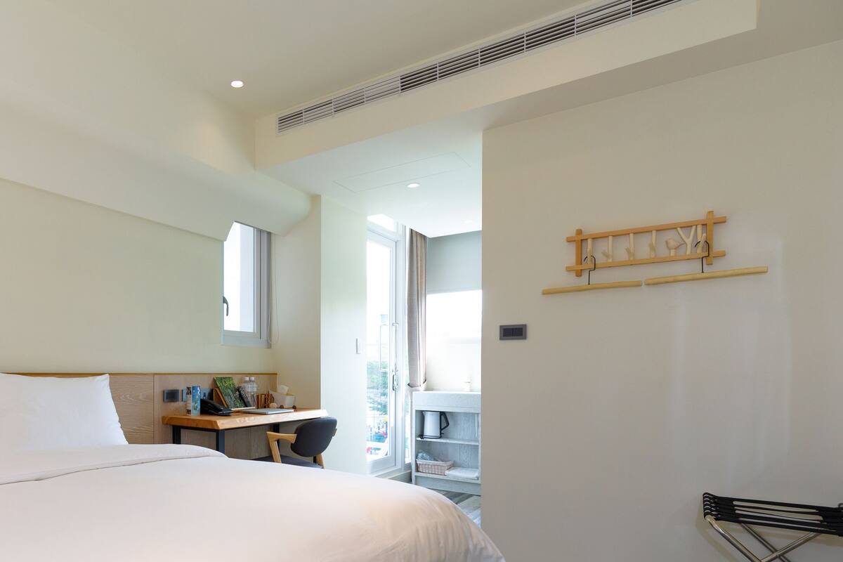Quarto de hotel com uma cama grande, uma escrivaninha com cadeira, uma mesinha e uma janela.