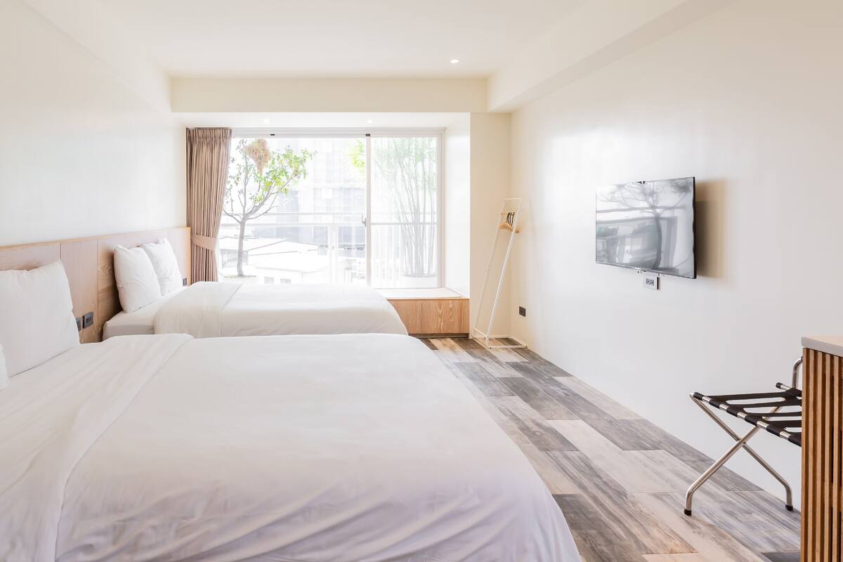 Quarto de hotel moderno com duas camas, uma janela grande, televisão de tela plana e piso de madeira.