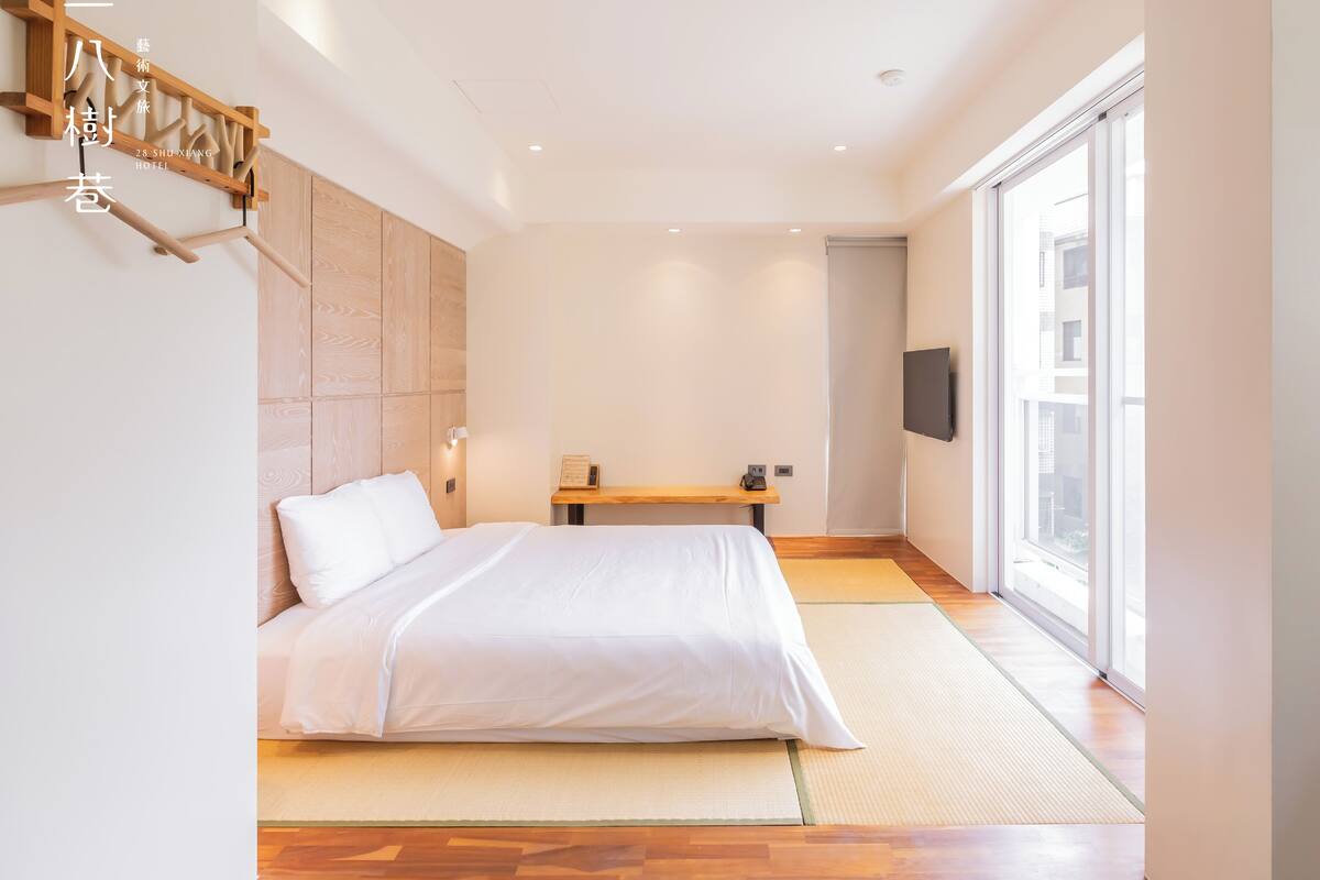 Quarto de hotel moderno com cama, cabeceira de madeira, mesa de cabeceira, TV de tela plana e uma janela grande.