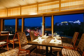 Uma sala de jantar com mesa e cadeiras de madeira, janelas amplas e vista para o horizonte urbano à noite.