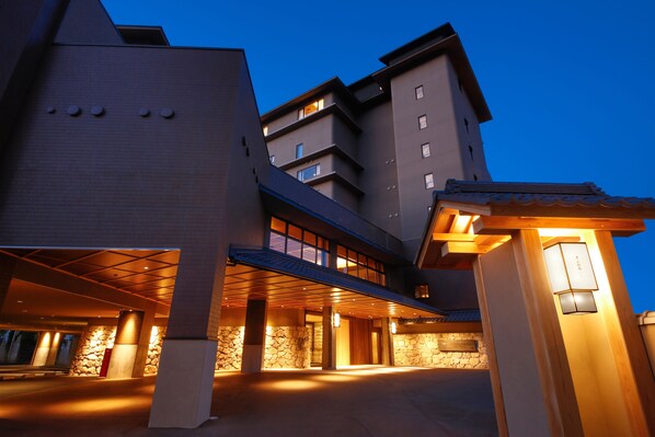 Uma entrada de hotel moderna com galeria coberta, pilares de pedra e fachada bem iluminada.
