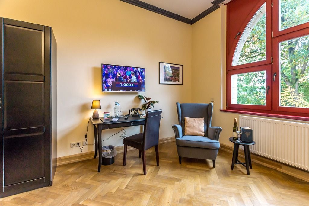 Um quarto com piso de madeira em padrão herringbone, uma escrivaninha com computador, uma cadeira, uma mesinha com uma garrafa e uma janela com moldura vermelha.