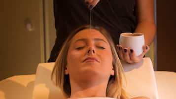 Uma pessoa recebendo tratamento facial, com uma toalha cobrindo o rosto e uma xícara na mão.