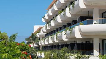 Um edifício moderno com várias varandas, algumas com plantas, e um caminho ladeado por plantas e flores tropicais.