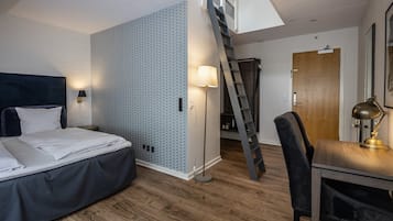 Quarto de hotel com uma cama, uma escrivaninha com abajur, uma escada e uma porta.