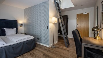 Quarto de hotel com uma cama, uma escrivaninha com abajur, uma escada e uma porta.