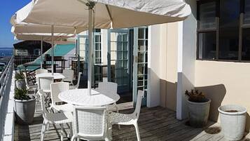 Um terraço com mobiliário de vime branco, incluindo mesas e cadeiras redondas, sob um grande guarda-sol branco.