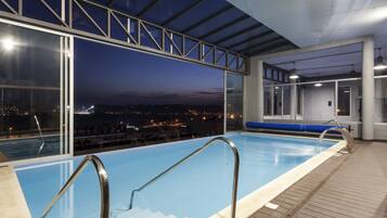Uma piscina interior com paredes de vidro transparente, guarda-corpo de metal e vista para a cidade ao entardecer.