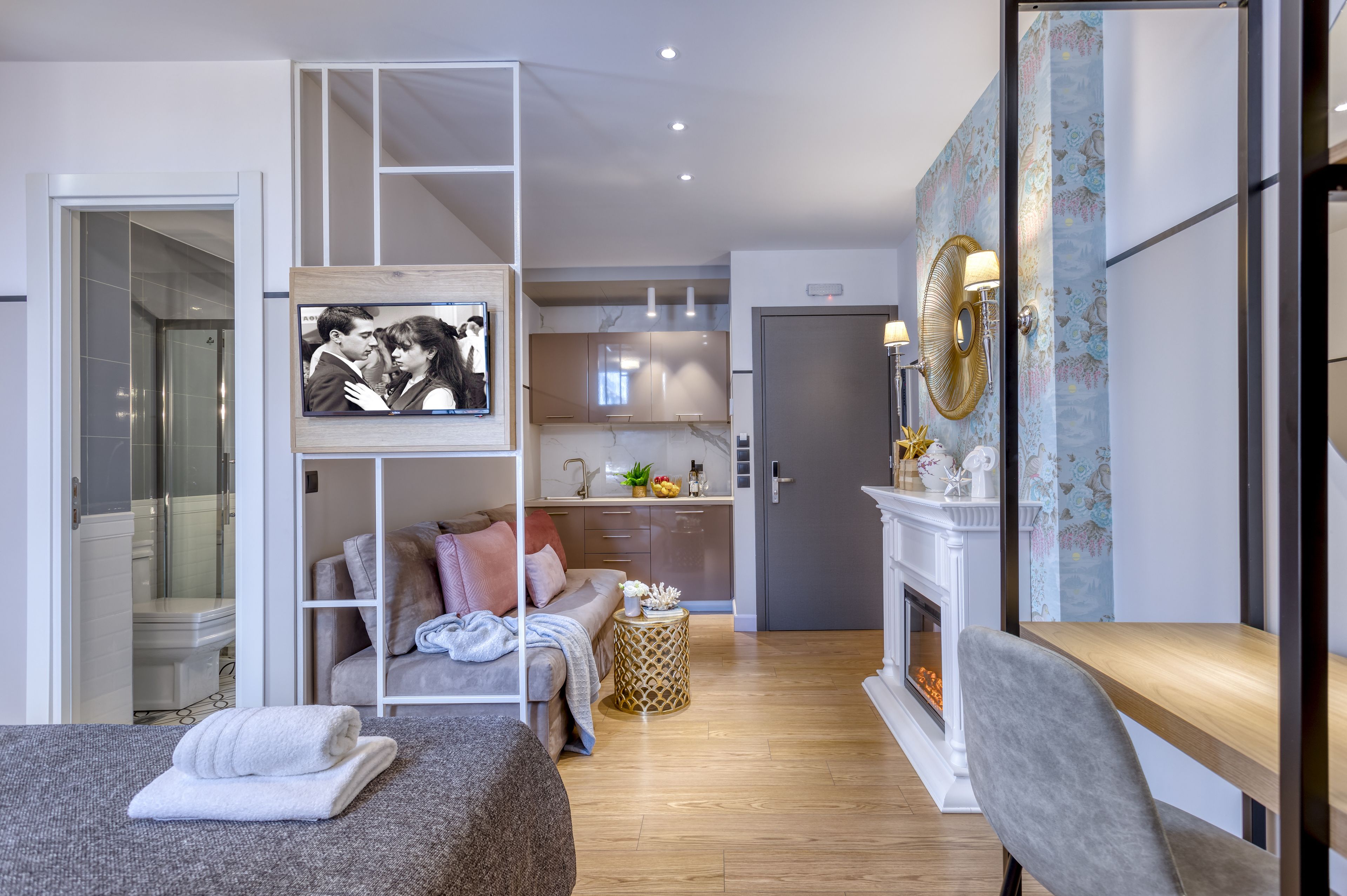 Quarto de hotel moderno com sofá, uma pequena mesa redonda, televisão fixada em parede branca, lareira e área de cozinha visível por uma porta aberta.