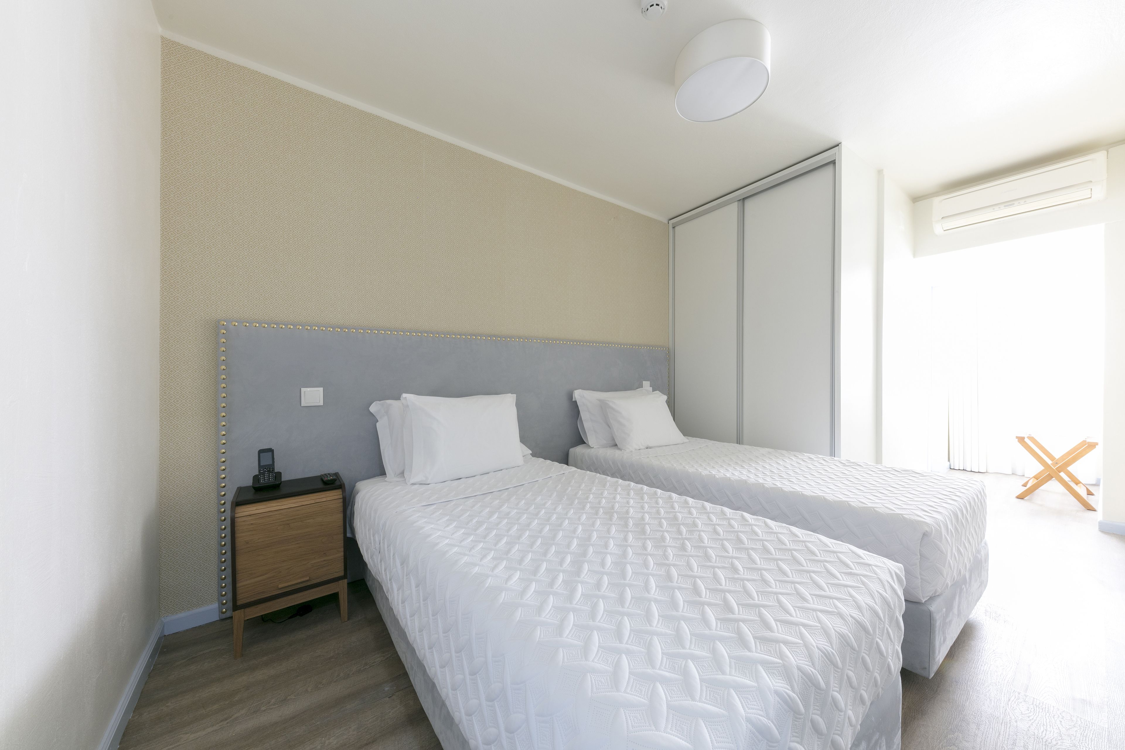 Quarto com duas camas, mesa de cabeceira, guarda-roupa e janela com persianas.