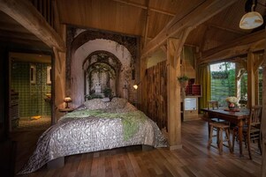 Quarto rústico com uma cama grande, vigas de madeira, um arco de pedra e um espaço de refeições com mesa e cadeiras.