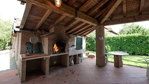 Forno de tijolos ao ar livre com fogo, uma pia e uma mesa sob uma estrutura de telhado de madeira.