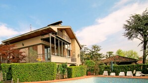 Uma casa moderna de dois andares com piscina, cercada por sebes e árvores bem cuidadas.