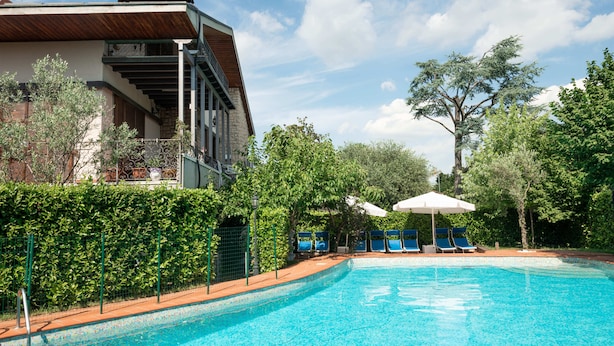 Piscina com espreguiçadeiras e guarda-sóis, cercada por vegetação e um edifício com varanda.