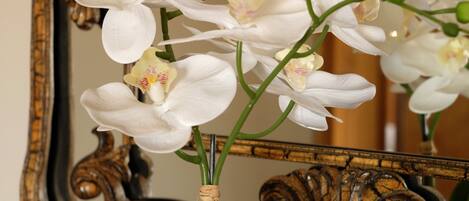 Close-up de orquídeas brancas com centros amarelos, amarradas a um caule verde, diante de um espelho de madeira com esculturas detalhadas.