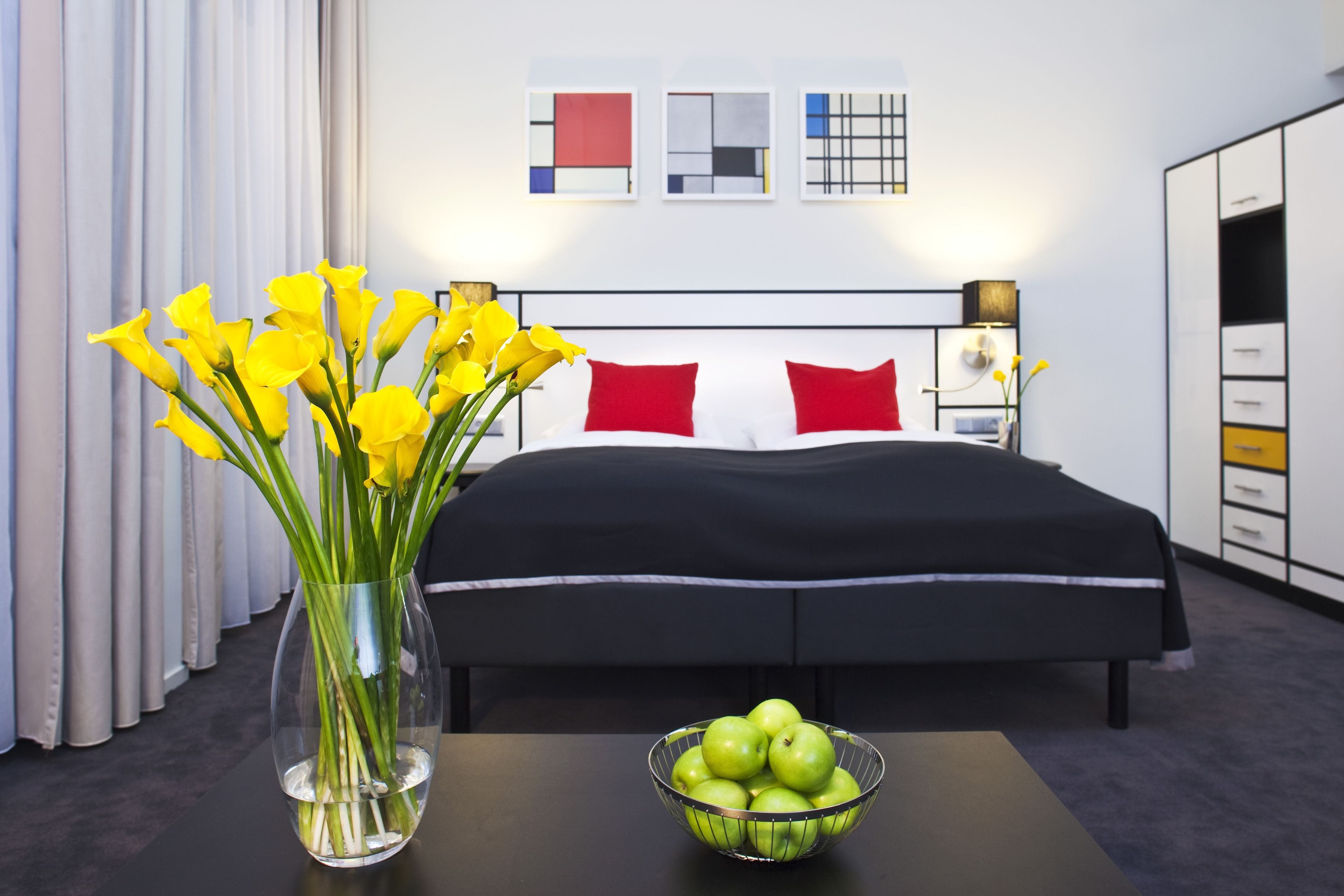 Quarto de hotel moderno com uma cama preta, almofadas vermelhas e brancas, um vaso com flores amarelas e uma tigela com maçãs verdes sobre uma mesa.