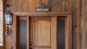 Uma porta dupla de madeira com uma placa que diz 'Welcome' acima, uma escultura de veado à direita e um candeeiro à esquerda.