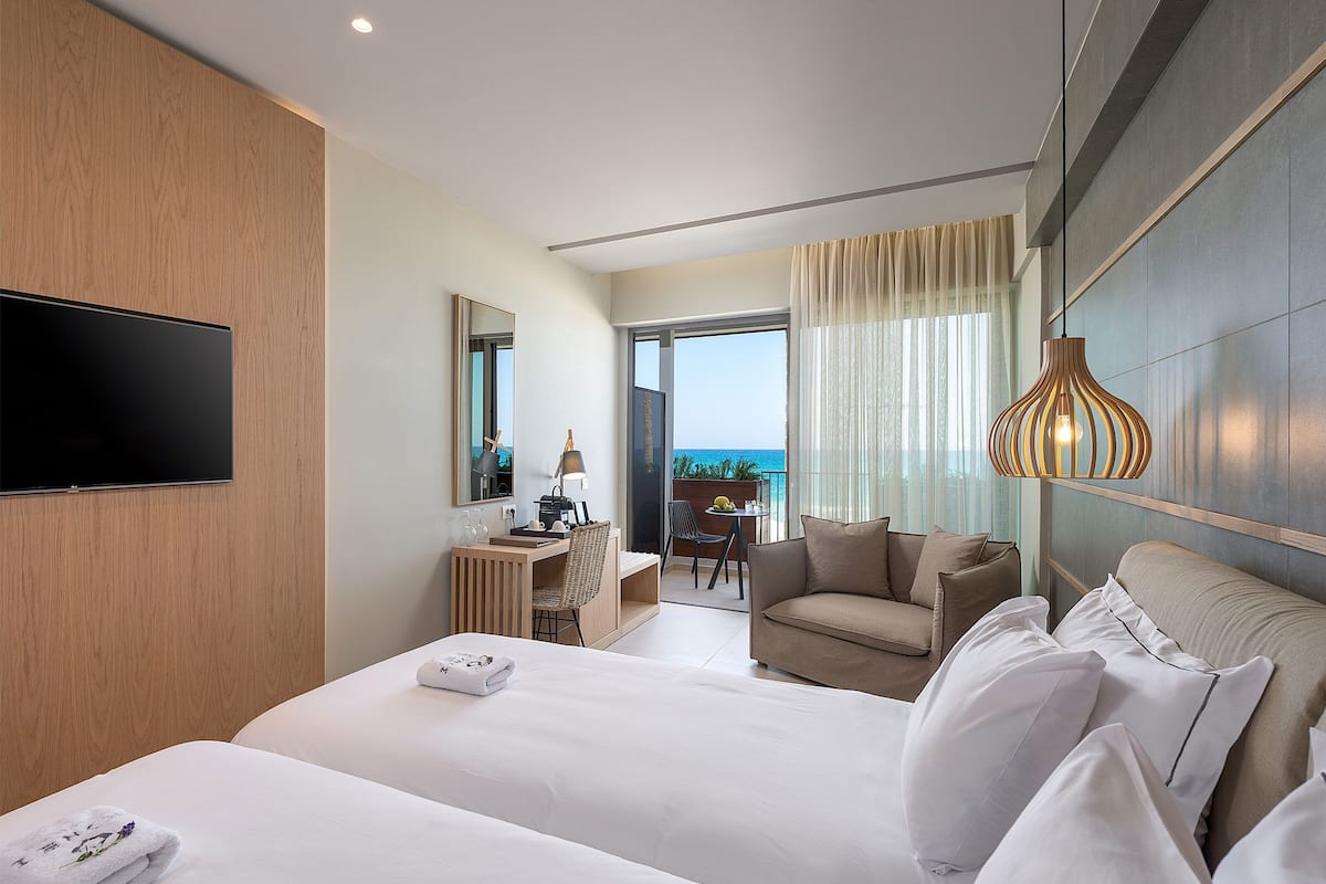 Quarto de hotel com uma cama grande, uma escrivaninha com cadeira, um sofá, televisão montada na parede e vista para o oceano pela janela.