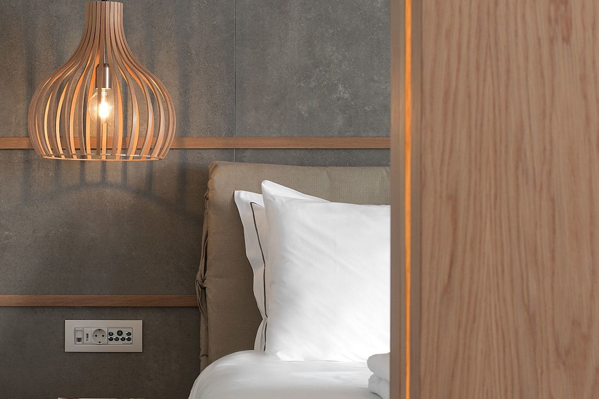 Quarto moderno com mesa de cabeceira de madeira, cama branca e luminária pendente diferenciada.