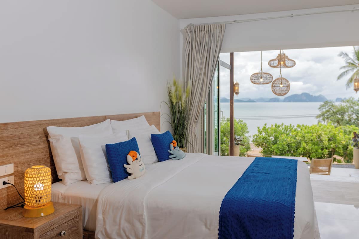 Quarto com uma cama grande, roupa de cama branca, mesa de cabeceira com abajur e vista para o mar através de uma varanda.