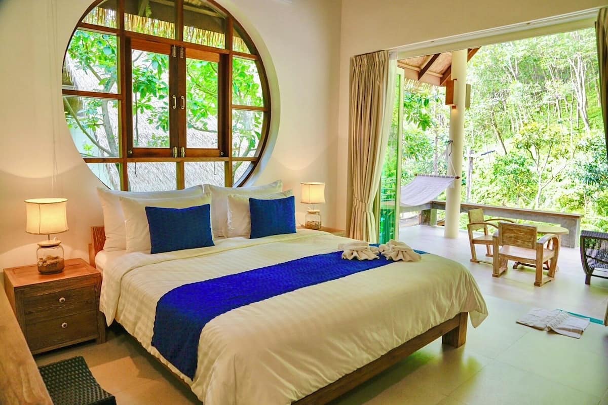 Quarto com uma janela ampla, cama com roupa de cama branca e azul, mesa de cabeceira com abajur e vista para o verde lá fora.