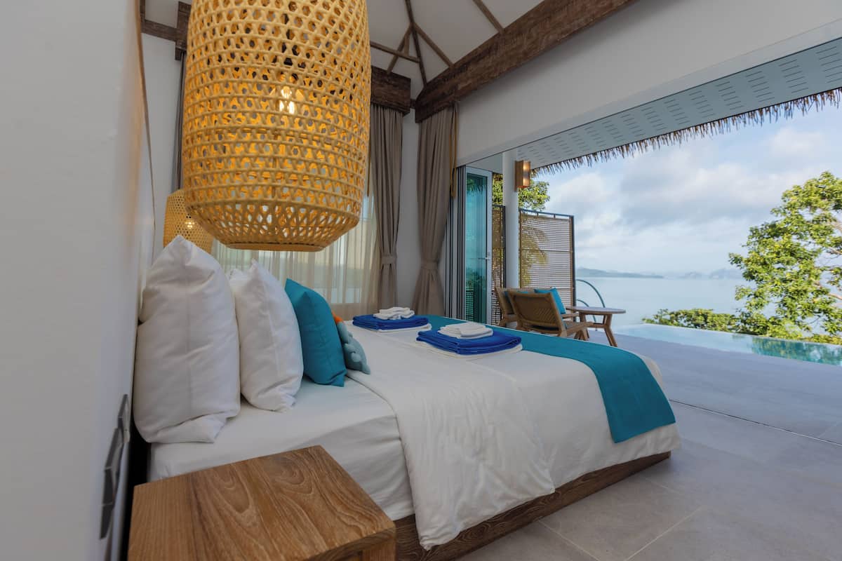 Quarto com cama, travesseiros, mesa de cabeceira, luminária pendente e vista para o mar.