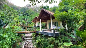 Um ecolodge isolado com estrutura de telhado de palha, uma ponte de madeira e vegetação exuberante.