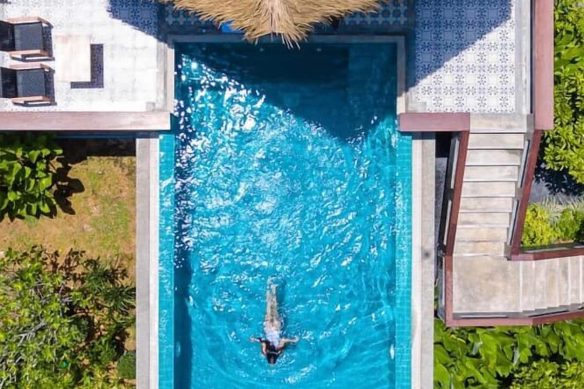 Vista aérea de uma piscina retangular com uma pessoa nadando, cercada por vegetação exuberante e uma estrutura com telhado de palha.