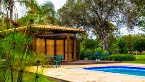 Um gazebo de madeira com telhado de azulejos, cercado por uma piscina e cadeiras verdes.