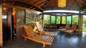 Uma área de estar em madeira com vários espreguiçadeiras, uma mesinha e um lustre no teto.