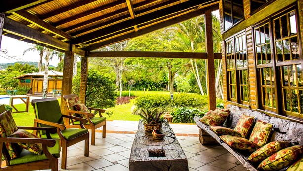 Uma área de estar coberta ao ar livre, com mobiliário de madeira, uma mesa central e vista para um jardim exuberante.